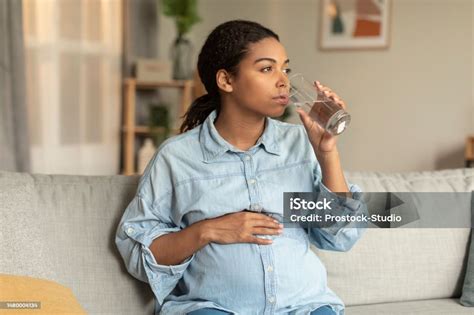 젊은 아프리카계 미국인 임산부는 큰 배꼽을 가지고 물 한 잔을 마시고 자유 시간을 즐기고 집에서 소파에 앉아 있다 25 29세에