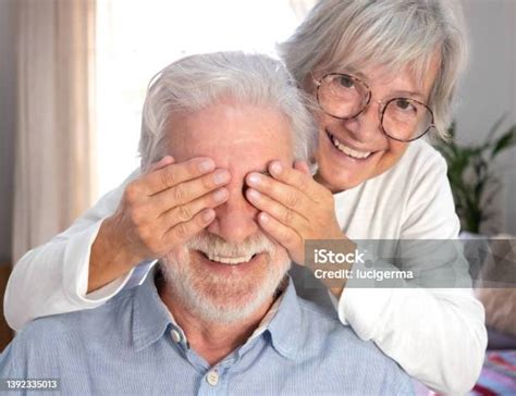 남편의 눈에 손을 얹고 카메라를 바라보는 아름다운 고위 백인 여성 집에있는 거실에서 장난스럽게 웃는 노인 부부 2명에 대한 스톡 사진 및 기타 이미지 Istock