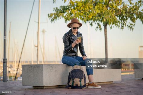 고급 모자 선글라스 가죽 재킷에 짧은 머리를 가진 30대 금발 여성이 바르셀로나의 요트 마린 옆 벤치에서 스마트폰을 사용하고 있다 30 39세에 대한 스톡 사진 및 기타