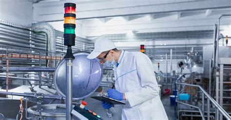 모자 마스크 및 흰색 코트의 기술자가 생산 공장에 서서 시트에 펜으로 메모를합니다 검사관은 낙농 공장에서 표시기를 제거합니다 엔지니어는 생산에 대한 통계를 유지합니다