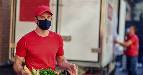 집에 계세요 검은 얼굴 보호 마스크를 쓴 젊은 백인 남성 택배는 식료품 상자를 들고 배경에 트럭이 있는 거리에 서 있습니다