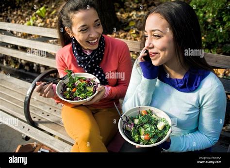Young Adult Female Twins Sitting On Park Bench Chatting On Smartphone And Eating Lunch Stock