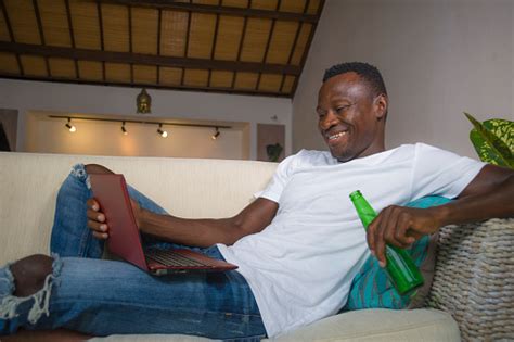 매력적이 고 행복 한 성공적인 검은 아프리카 미국 젊은이 인터넷 비즈니스 성공 개념에서 쾌활 한 마시는 맥주 병을 웃 고 거실 소파에서 노트북 컴퓨터와 네트워킹 30 39