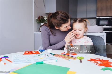 엄마와 4살짜리 딸이 하얀 테이블에 함께 앉아 Diy 공예품을 만들고 있습니다 귀여운 아이가 카드에 빨간 종이 하트를 조심스럽게 올려 놓고 엄마는 사랑의 미소를 지으며