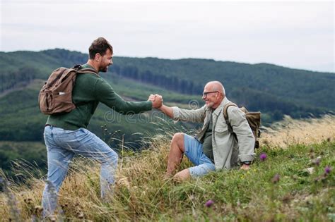 Adult Son Helping Older Father To Stand Up Hiking Togeter In Nature