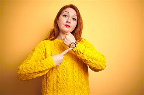 아름다운 빨간 머리 여자 착용 겨울 스웨터 위에 서 고립 된 노란색 배경 에 서 서둘러 가리키는 시계 시간 조바심 편안한 표정으로 카메라를 보고 가리키기에 대한 스톡 사진