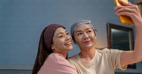 웃는 아시아 여자 친구들은 침대에 앉아 집에서 함께 얼굴에 스킨 케어 트리트먼트 페이셜 마스크를 바르고 있다 스마트폰을 사용하여 행복과 함께 셀카를 찍는 여성 게이 커플