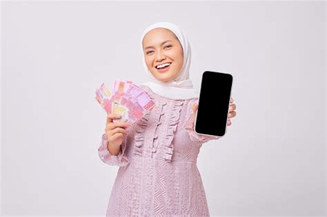 히잡과 보라색 드레스를 입은 쾌활한 아름다운 아시아 무슬림 여성이 빈 화면과 흰색 스튜디오 배경에 격리된 돈 지폐가 있는 휴대폰을 들고 있다 Eid Ul Fitr에 대한