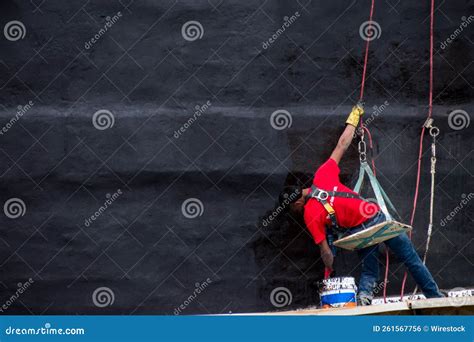 Man Sitting On A Wooden Suspended Scaffold And Painting A Wall In The Color Black Stock Photo