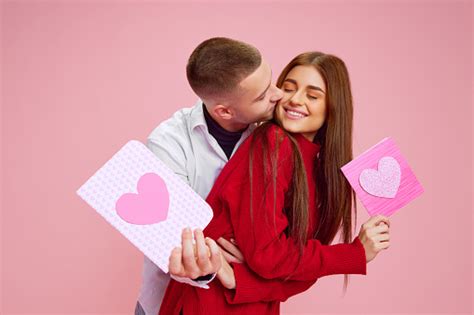 아름 다운 젊은 커플 남자와 여자가 포옹 미소 분홍색 스튜디오 배경에 하트와 함께 엽서를 들고 있습니다 키스 발렌타인 데이 인연 18 19세에 대한 스톡 사진 및 기타