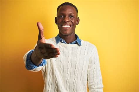 데님 셔츠와 흰색 스웨터를 입은 아프리카 계 미국인 남성이 고립 된 노란색 배경 위에 인사와 환영으로 악수를 제공합니다 성공적인 비즈니스 겨울에 대한 스톡 사진 및 기타