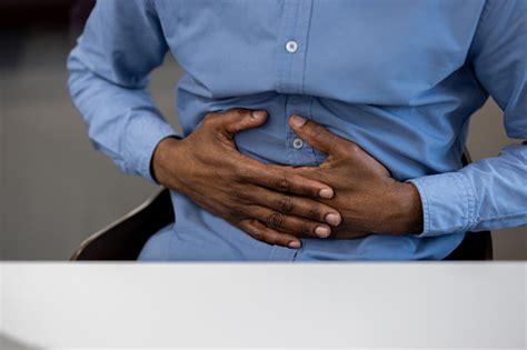 책상에 앉아서 불편함을 겪고 있는 성숙한 아프리카계 미국인 사업가 그는 배를 잡고 있는데 이는 통증이나 소화 불량일 수 있음을 나타냅니다 비즈니스 복장과 고요한 분위기는