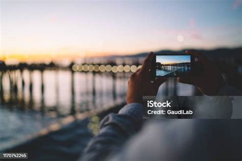 현대 스마트 폰 카메라를 사용하여 자연 환경의 저녁 황혼에 사진을 찍는 남자의 자른 이미지 황혼에 응용 프로그램을 통해 바다 물의 사진을 만드는 휴대 전화를 들고 힙스터