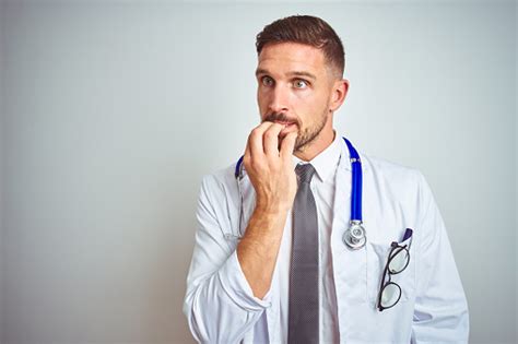 젊은 잘 생긴 의사 남자 흰색 전문 코트를 입고 고립 된 배경 에 찾고 스트레스와 긴장 한 입 물고 손톱에 손으로 불안 문제