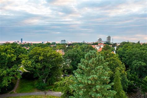 Urban Skyline With Green Trees In Foreground Under Cloudy Sky At Dusk
