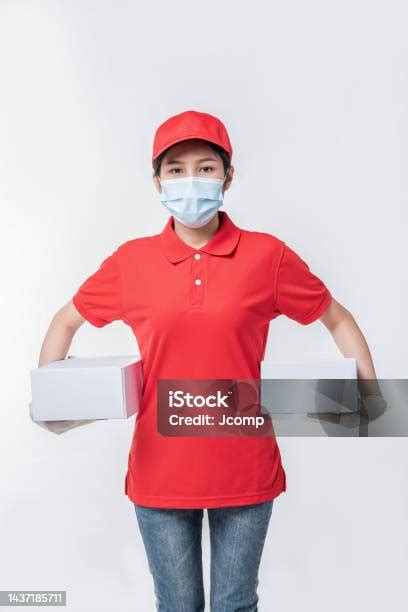 빨간 모자 빈 티셔츠 유니폼 얼굴 마스크 장갑을 낀 의식이 있는 젊은 배달원의 이미지는 밝은 회색 배경 스튜디오에 격리된 빈 흰색 판지 상자 팬데믹 코로나바이러스 바이러스