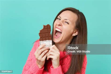 손에 들고와 초콜릿 바 물고 뜬된 분홍색 스웨터에 미친 쾌활 한 젊은 여자 블루 터키석 벽 배경 스튜디오 초상화에 고립 사람들의 라이프 스타일 개념입니다 복사 공간을 비웃는