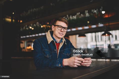 세련된 커피숍에서 휴식을 취하며 스마트폰으로 소셜 네트워크에서 프로필을 업데이트하는 동안 카메라에 미소 짓는 데님 재킷을 입은