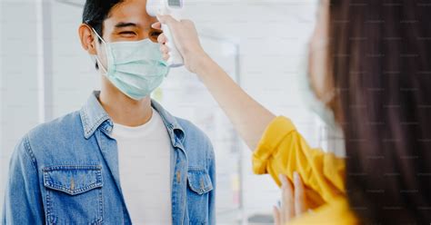 아시아 여성 접수 원은 사무실에 들어가기 전에 적외선 온도계 검사기 또는 온도 총을 고객의 이마에 착용 보호 안면 마스크를 사용합니다 코로나 바이러스 이후의 라이프 스타일