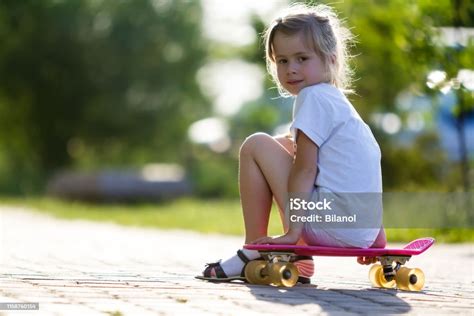 흰색 반바지와 티셔츠에 꽤 귀여운 작은 금발 소녀는 분홍색 스케이트 보드에 앉아 밝은 흐린 여름 배경에 카메라에 미소 어린이 활동과 활동적인 라이프 스타일 개념 감정에 대한