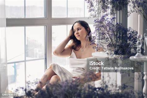 가벼운 실크 드레스를 입은 아름다운 젊은 장발 여성이 2022 년의 석고 필라 트렌디 한 색상으로 장식 된 방의 탁 트인 창문을 바라 봅니다 2022년에 대한 스톡 사진 및