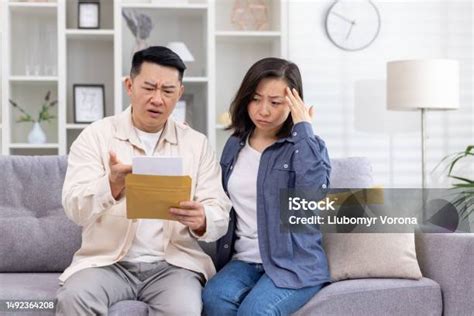 화가 난 아시아 가족 소파에 거실에 함께 앉아 있는 남자와 여자 부부는 나쁜 소식이 담긴 편지 메일 봉투 알림을 받았고 받은 메시지에 불만이 있었다 양 부모에 대한 스톡