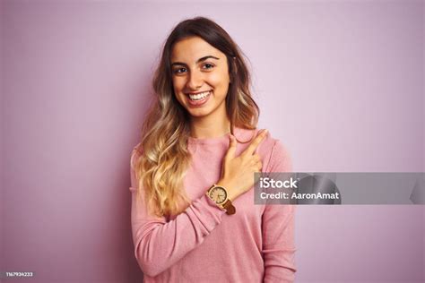 분홍색 고립 된 배경에 스웨터를 입고 있는 젊은 아름다운 여성은 얼굴에 행복하고 자연스러운 표정으로 손과 손가락으로 가리키는 얼굴의 미소와 함께 쾌활합니다 가리키기에 대한