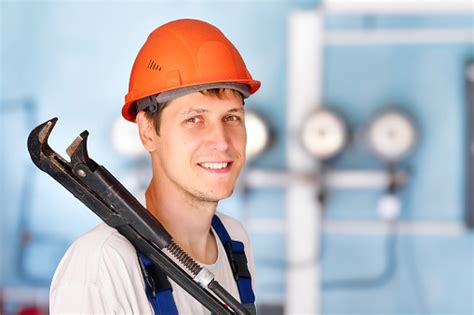 긍정적 인 산업 배관공 또는 헬멧을 쓴 작업자는 어깨에 큰 조절 렌치를 들고 미소를 짓습니다 흐릿한 센서와 압력 게이지의 배경에 대한 실내 인물 사진 연관공에 대한 스톡