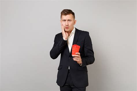 커피 또는 차 회색 배경에 고립 된 종이 컵을 들고 뺨에 손을 넣어 고전적인 검은 양복에 충격된 젊은 비즈니스 사람 성과 경력 부 비즈니스 개념입니다 복사 공간을 비웃는 다