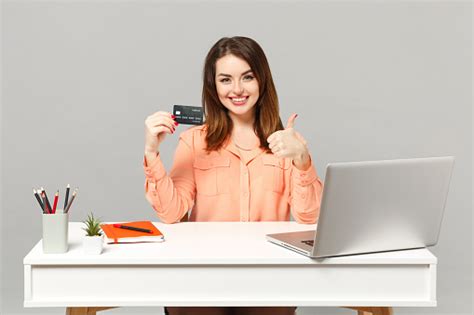 회색 배경에 고립 된 Pc 노트북과 책상에서 신용 카드 작업을 유지 엄지손가락을 보여주는 파스텔 옷에 젊은 웃는 여자 성취