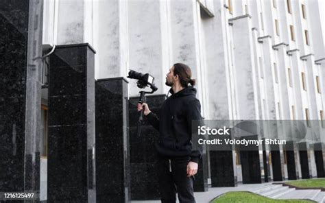 3축 짐벌 안정기에서 전문 카메라를 들고 있는 젊은 전문 비디오 그래퍼 프로 장비는 흔들리지 않고 높은 품질의 비디오를 만드는 데 도움이됩니다 검은 후드를 입고 카메라맨은