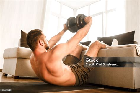 소파에 집에서 아령과 함께 운동 복근을하고 젊은 남자 자신의 아파트에서 운동 활동에 하드 워킹 티셔츠없는 남자 스포츠맨의 보기를 잘라 바닥에 누워 있는 사진에 아령 남자에