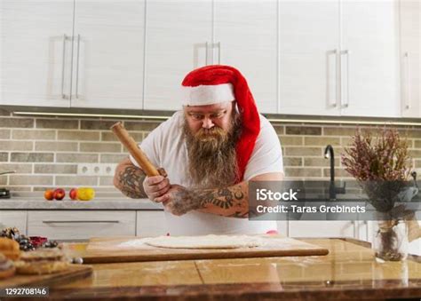 산타 클로스 모자를 쓴 좌절하고 화가 난 수염을 기리는 남자가 야구 방망이처럼 나무 롤링 핀을 들고 반죽을 바라본다 가정 생활에 대한 스톡 사진 및 기타 이미지 Istock