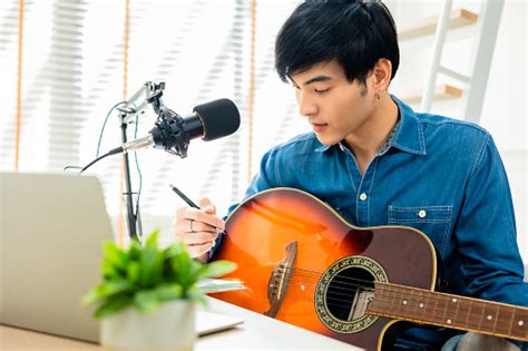 아시아 청년 작곡가는 어쿠스틱 기타를 연주하고 스마트 폰에서 노래를 듣고 생각 하 고 종이 가사 노래 홈 스튜디오의 거실에 앉아 쓰기 합니다 가정에서의 음악 제작 개념