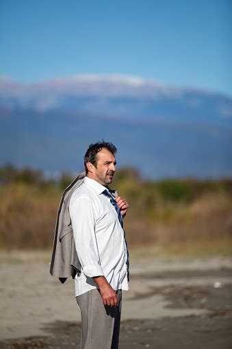 흰색 셔츠 블루 넥타이 롤업 바지와 그의 어깨에 코트를 가진 중 년 기업 남자 올림푸스 산을 배경으로 해변에 맨발로 탄 그리스 올림푸스 산에 대한 스톡 사진 및 기타 이미지