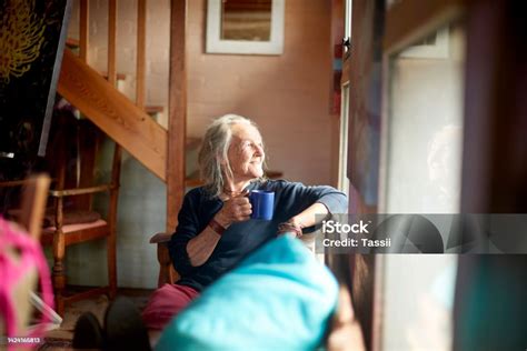 노인 여성은 아침에 거실에서 커피 한 잔과 함께 휴식을 취하고 미소를 짓습니다 행복한 노인 여성이 따뜻한 차를 마시고 아파트의 창문 전망을보고 생각하고 은퇴를 즐깁니다 65