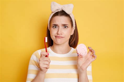 마시멜로와 칫솔을 보여주고 노란색 배경 위에 고립된 줄무늬 티셔츠를 입고 불확실한 표정 구강 건강 또는 설탕 마시멜로로 카메라를 바라보는 어리둥절한 여성 20 24세에 대한