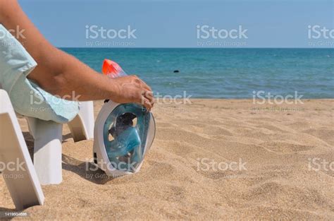 한 남자가 파도와 안개로부터 보호와 함께 스쿠버 다이빙을위한 마스크를 손에 들고 바다 근처의 모래 사장에있는 하얀 일광욕 용 의자에 누워 있습니다 남자에 대한 스톡 사진 및