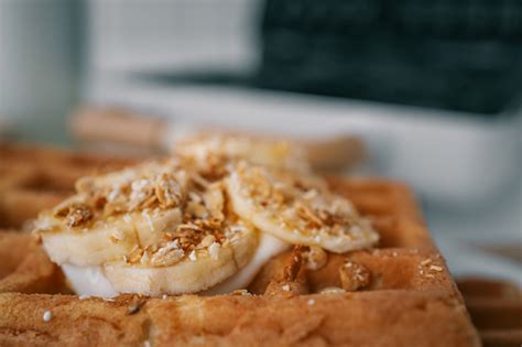 바나나 슬라이스 요구르트 그래놀라를 곁들인 갓 구운 와플을 화이트 플레이트에 담았습니다 흐릿한 배경에 놓인 차 한 잔과 와플 다리미는 따뜻하고 매력적인 아침 식사 환경을