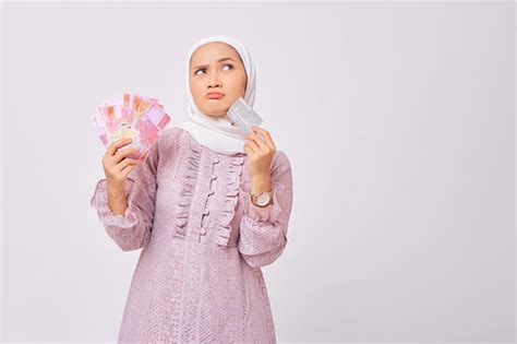히잡과 보라색 드레스를 입고 흰색 스튜디오 배경에 격리된 신용 카드와 돈 지폐를 들고 있는 잠겨 있는 젊은 아시아 무슬림 여성 Eid Ul Fitr에 대한 스톡 사진 및