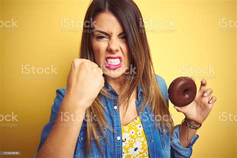 노란색 배경에 초콜릿 도넛을 먹는 젊은 아름다운 여자는 화가 좌절 분노와 함께 소리 미친 고함 제기 손으로 고함 분노 개념 도넛에 대한 스톡 사진 및 기타 이미지 Istock