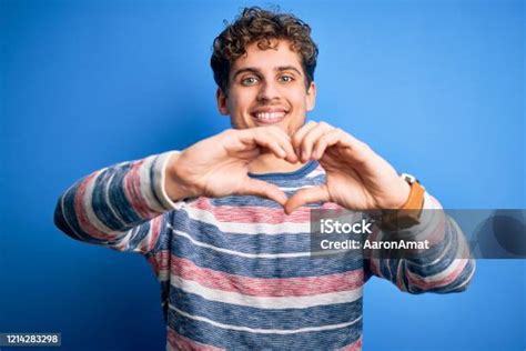 젊은 금발 잘생긴 남자 곱슬 머리 를 입고 줄무늬 스웨터 위에 파란색 배경 미소 사랑 보여주는 심장 기호와 손으로 모양 로맨틱 한 개념 개념에 대한 스톡 사진 및 기타