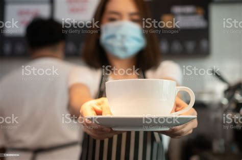 커피컵의 선택적 초점과 전염병을 예방하고 커피숍의 고객에게 제공하기 위해 얼굴 마스크를 착용한 아시아 바리스타 여성의 배경